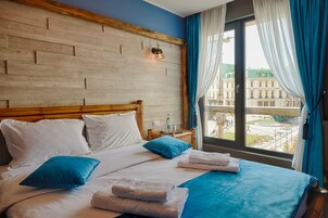 Quarto de hotel com cabeceira de madeira, cama grande com roupa de cama branca e azul, mesa de cabeceira com garrafa de água e vista para edifícios pela janela.