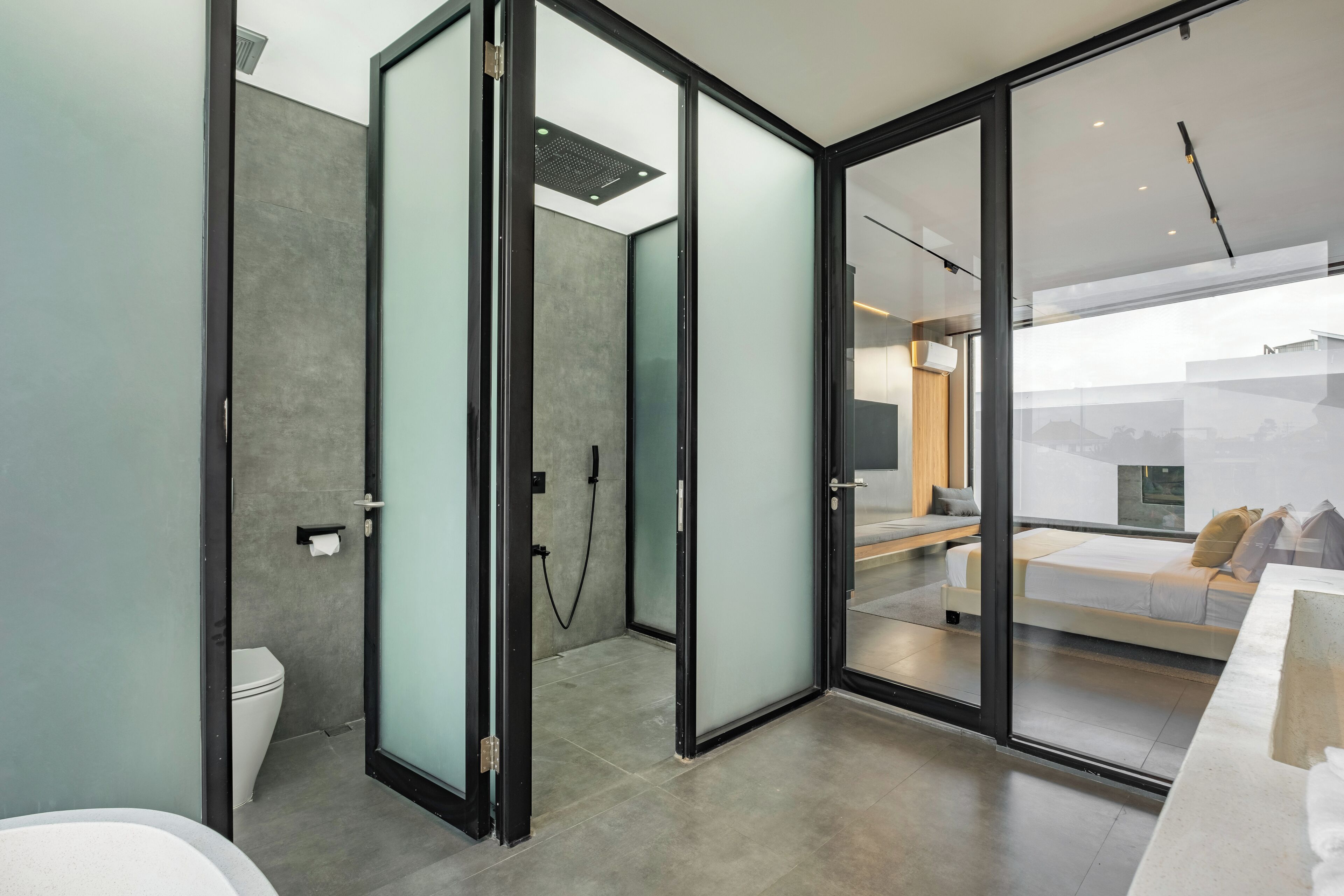Um banheiro moderno com vaso sanitário, chuveiro e portas de vidro que dão para um quarto com cama e sofá.
