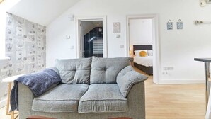 Uma sala de estar moderna com um sofá cinzento, uma mesa pequena e uma escadaria que leva a um quarto.
