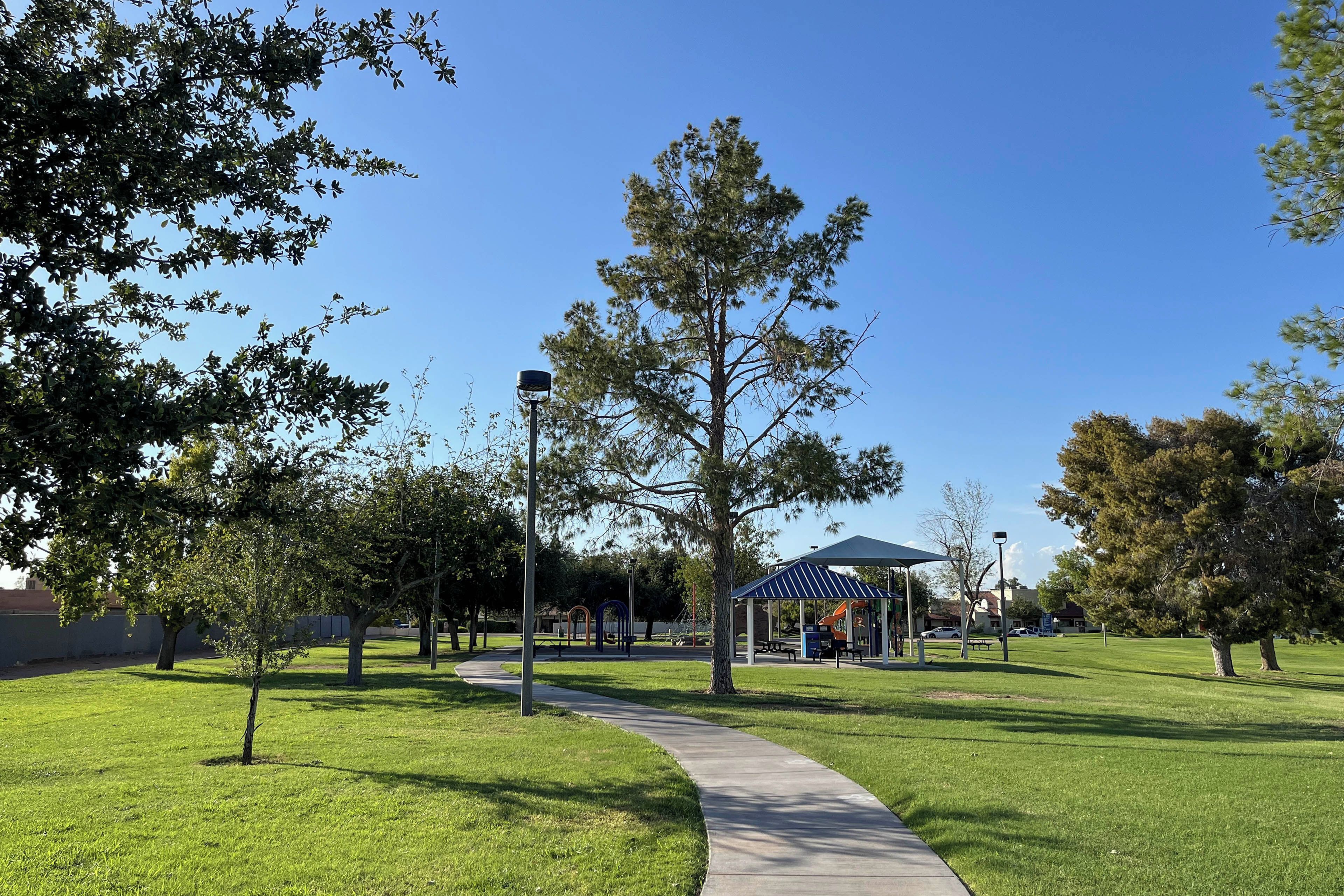 Um parque com calçada pavimentada, árvores e área de recreação coberta.