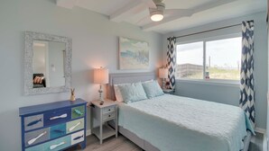 Quarto com cama, mesas de cabeceira, cômoda, ventilador de teto, janela com cortinas e uma pintura com tema de praia.