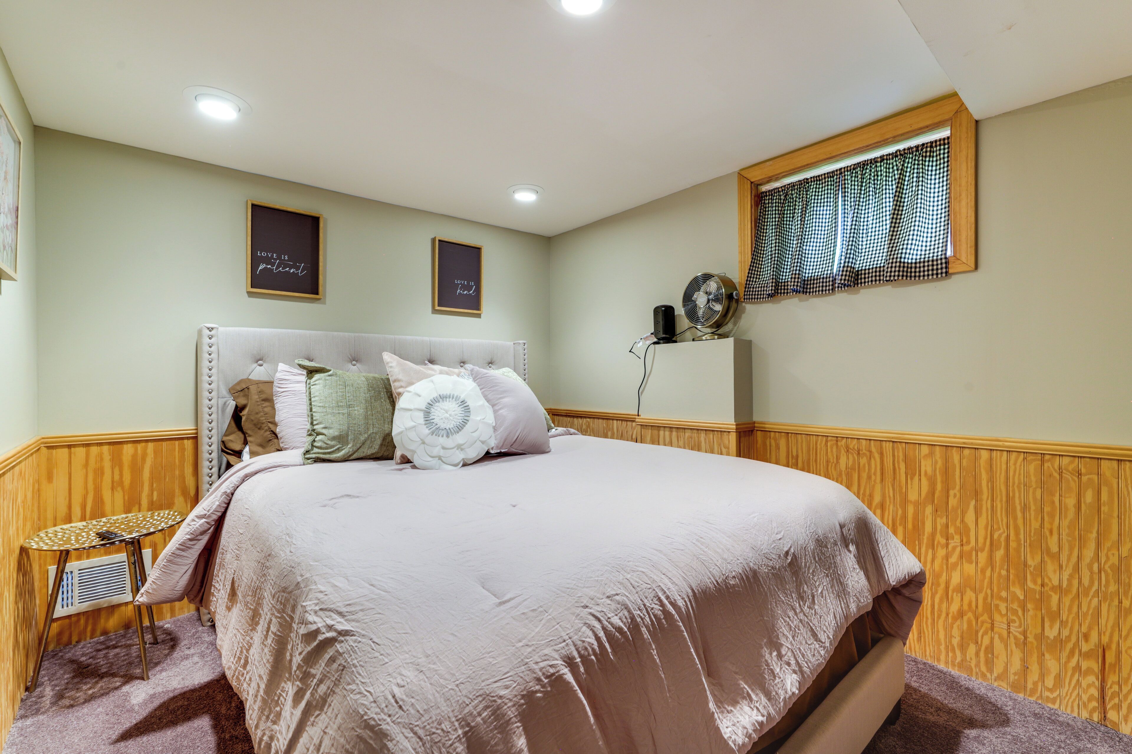 Quarto bem arrumado com uma cama grande, uma mesinha de cabeceira, um ventilador fixado na parede e duas frases inspiradoras emolduradas.
