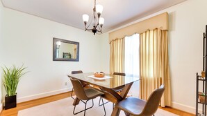 Uma sala de jantar com mesa de vidro, quatro cadeiras, lustre, planta em vaso, espelho e cortinas.