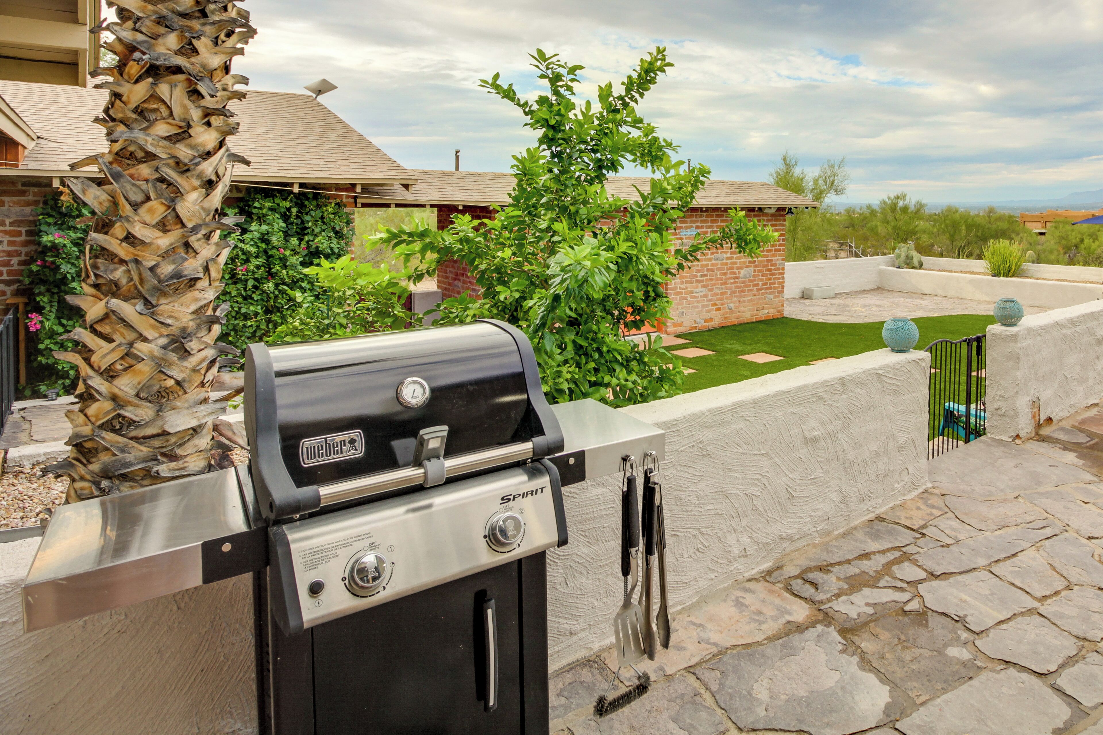 Uma churrasqueira Weber Spirit com um fundo de parede de pedra, uma palmeira e vista para uma casa de tijolos com gramado.