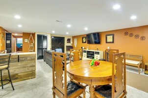 Uma sala de jantar com mesa e cadeiras de madeira, cozinha ao fundo e TV fixada na parede.