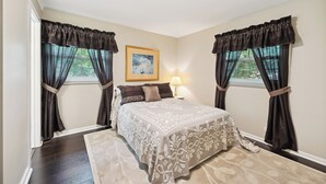 Quarto com cama, duas janelas com cortinas, um quadro na parede e uma mesa de cabeceira com abajur.