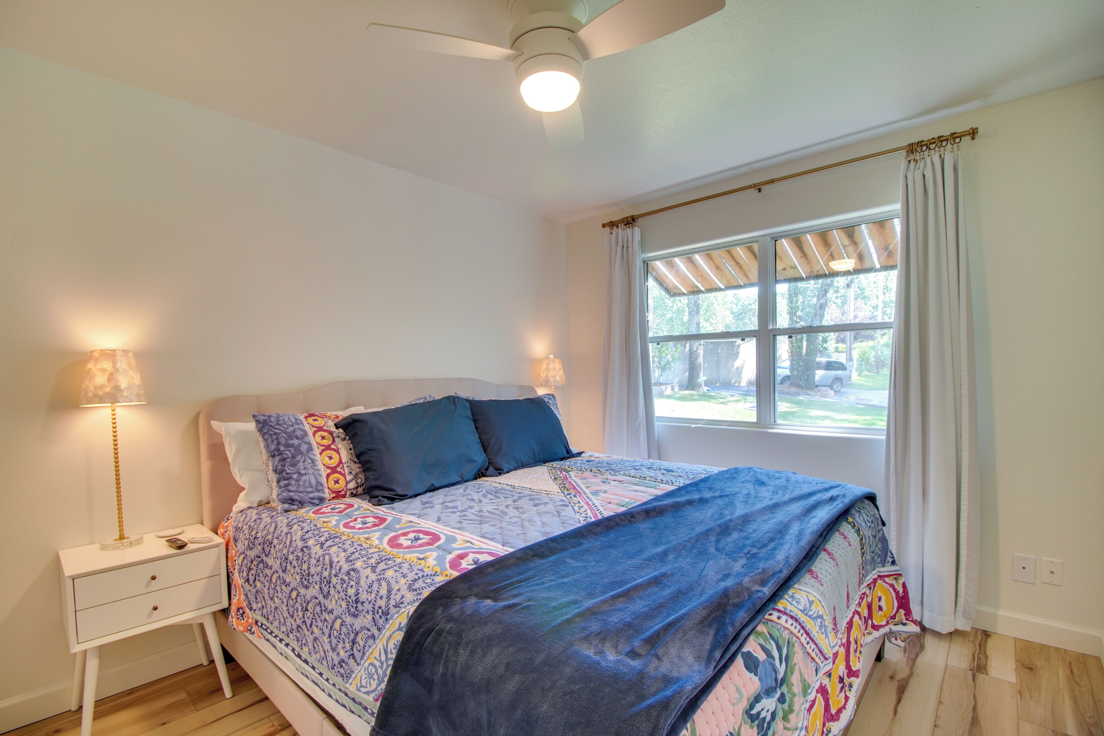 Quarto com cama, mesas de cabeceira, ventoinha no teto, janela com cortinas e vista para o exterior.