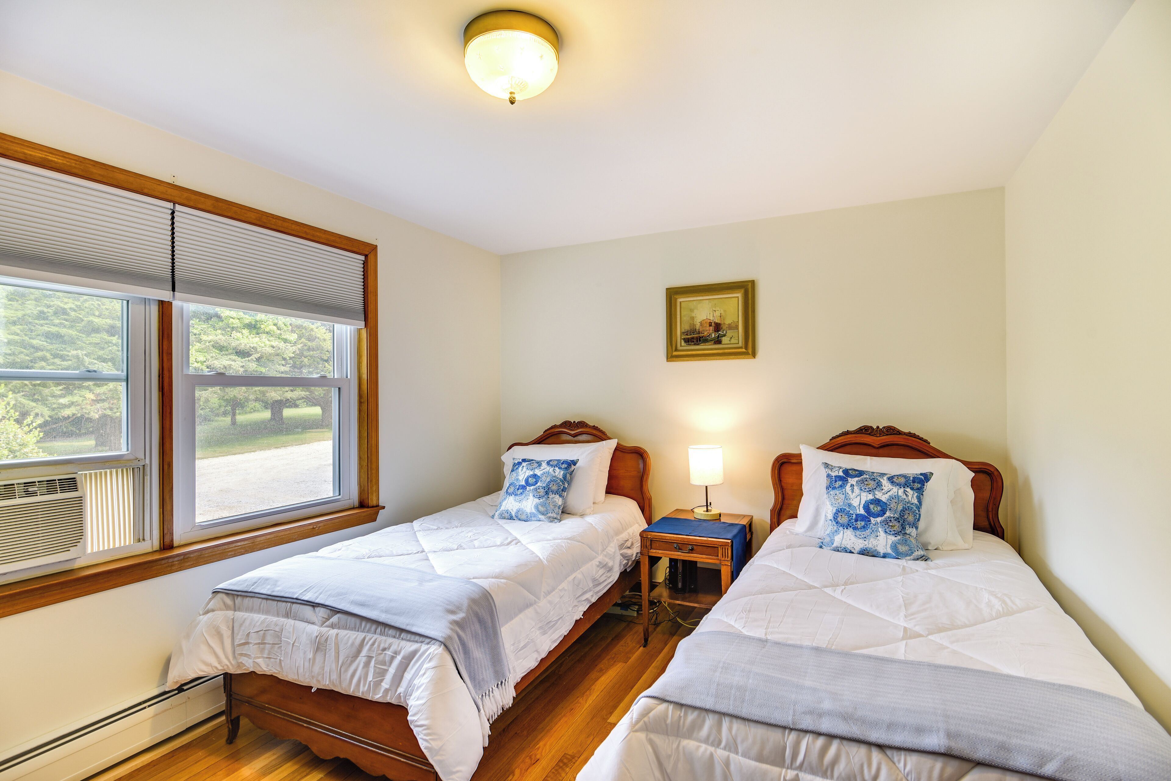 Quarto com duas camas, mesa de cabeceira com luminária, janela com persianas e quadro na parede.