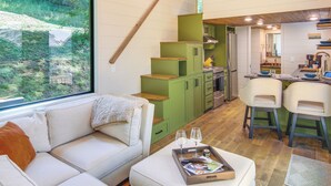 Interior moderno com sofá, área de refeições com duas cadeiras, cozinha com armários verdes e uma grande janela com vista para vegetação.