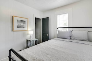 Um quarto com uma cama, um quadro na parede, uma mesinha de cabeceira com uma luminária e uma porta.