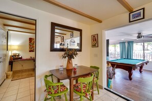 Uma sala de jantar com uma mesa de madeira, cadeiras verdes, um vaso com flores e um bilhar ao fundo.