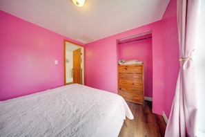 Quarto com paredes cor-de-rosa, cómoda de madeira e cama com colcha branca.