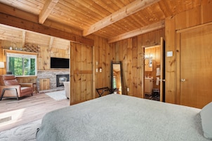 Interior de uma cabana de madeira com lareira, TV, cama e banheiro visível através de uma porta aberta.