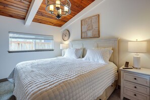 Quarto com cama grande, teto de madeira, relógio de parede, mesa de cabeceira com abajur e janela com persianas.