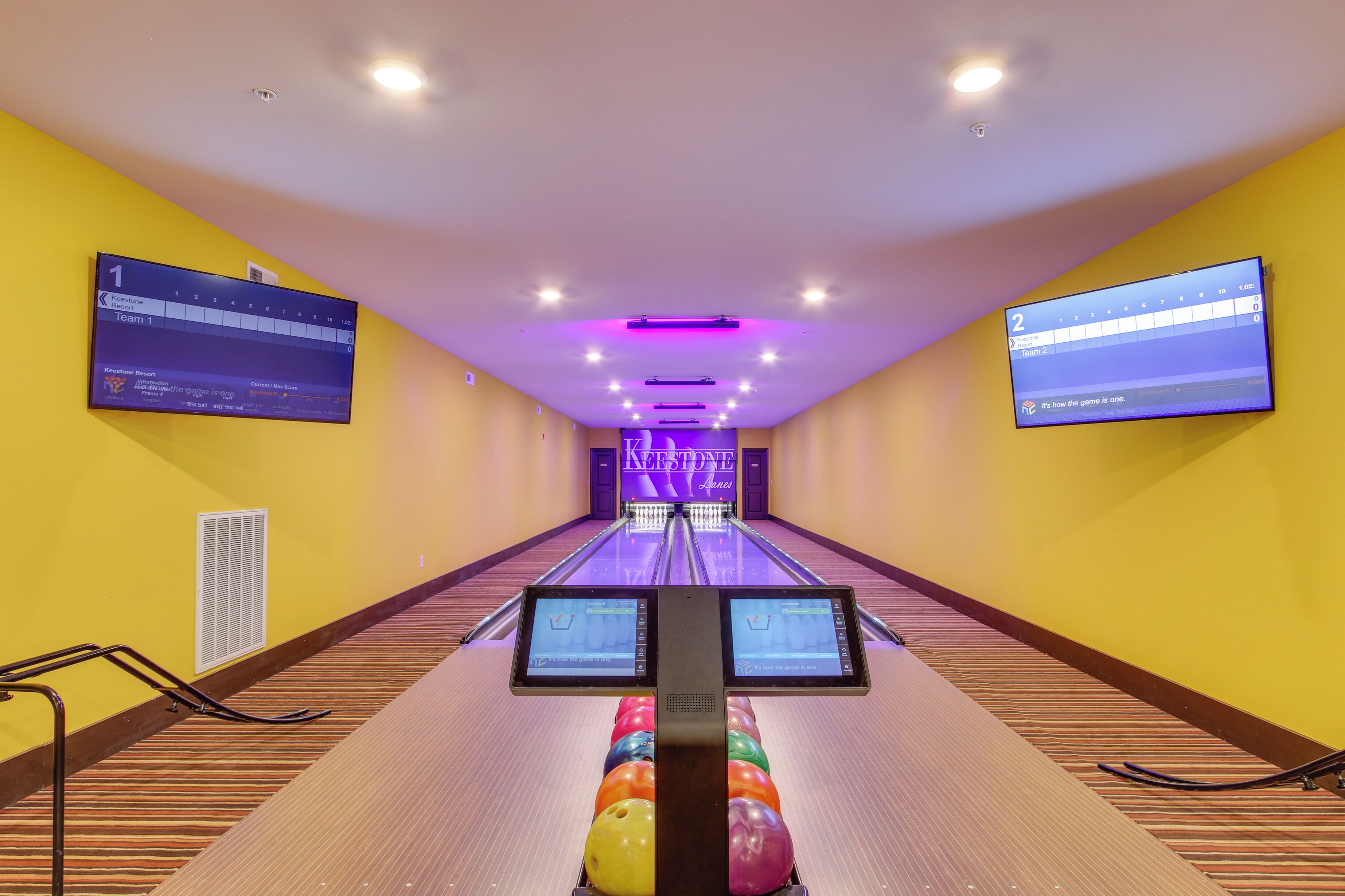 Uma pista de boliche com duas telas mostrando placares, uma pista de bowling e iluminação roxa ao fundo.