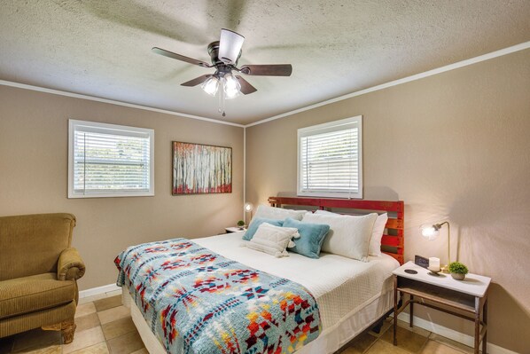Um quarto com uma cama, uma cadeira, uma mesa de cabeceira, um ventilador de teto e duas janelas com persianas.