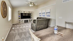 Uma sala de estar com uma grande TV de tela plana, um sofá seccional e uma mesa de jantar com uma garrafa de água e uma caixa em cima.