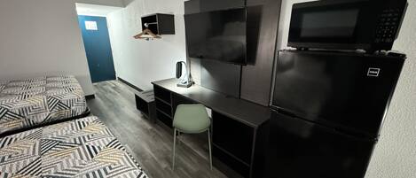 Quarto compacto com duas camas estampadas, uma escrivaninha com cadeira, um frigorífico preto e uma TV na parede.