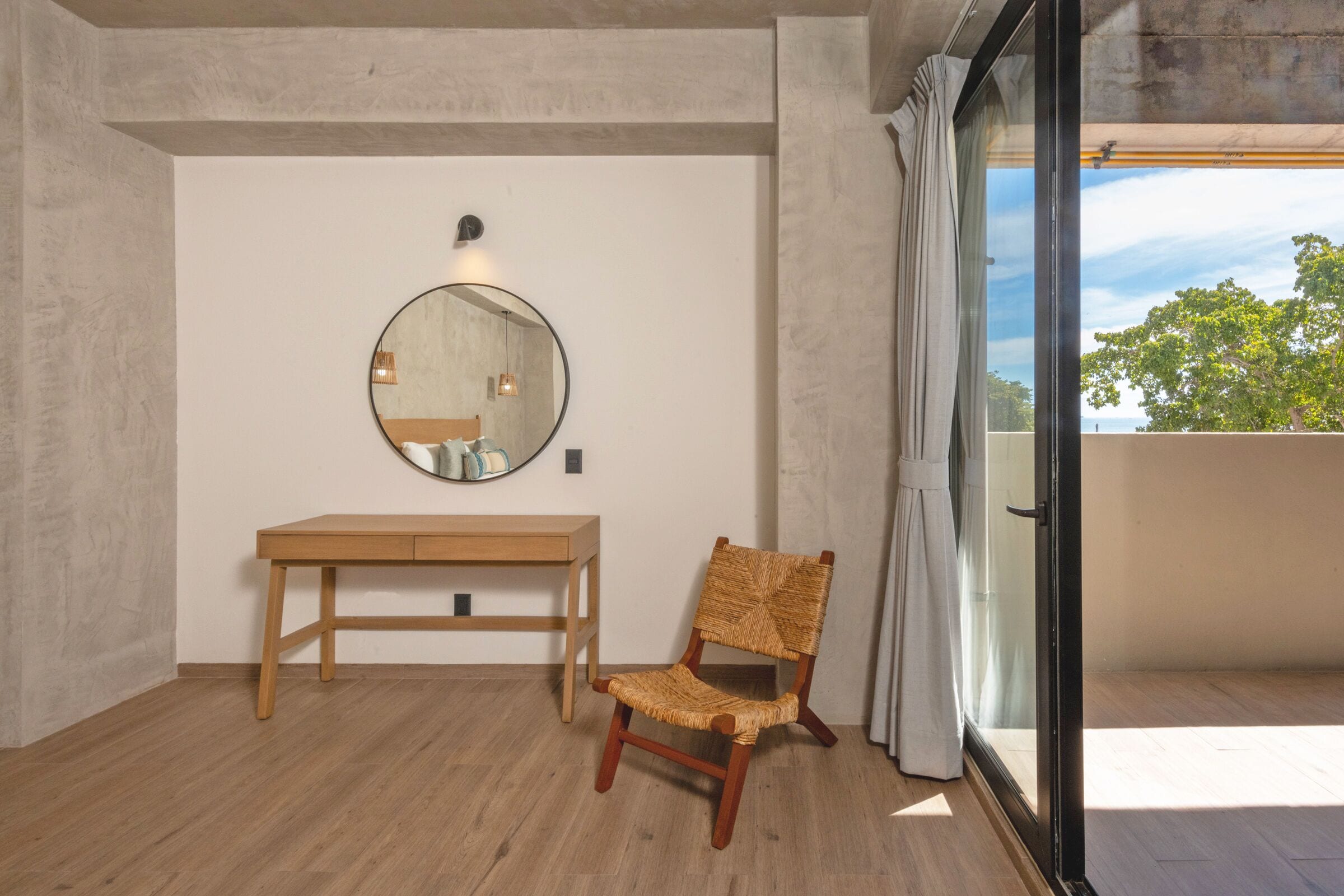 Quarto com escrivaninha de madeira, cadeira de vime, espelho redondo na parede e uma janela grande com vista para árvores.