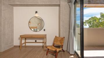 Quarto com escrivaninha de madeira, cadeira de vime, espelho redondo na parede e uma janela grande com vista para árvores.