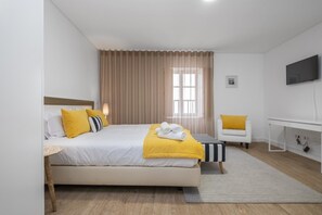 Quarto de hotel moderno com uma cama grande, uma mesa de cabeceira, uma cadeira, uma escrivaninha e uma TV fixada na parede.