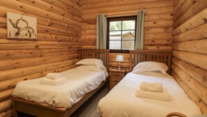 Quarto de cabana de tronco com duas camas de madeira, uma mesinha, um abajur e uma janela com cortinas.