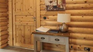 Interior de uma cabana de madeira com porta de madeira, uma mesa com abajur, um caderno e um quadro na parede com garrafas de vinho.