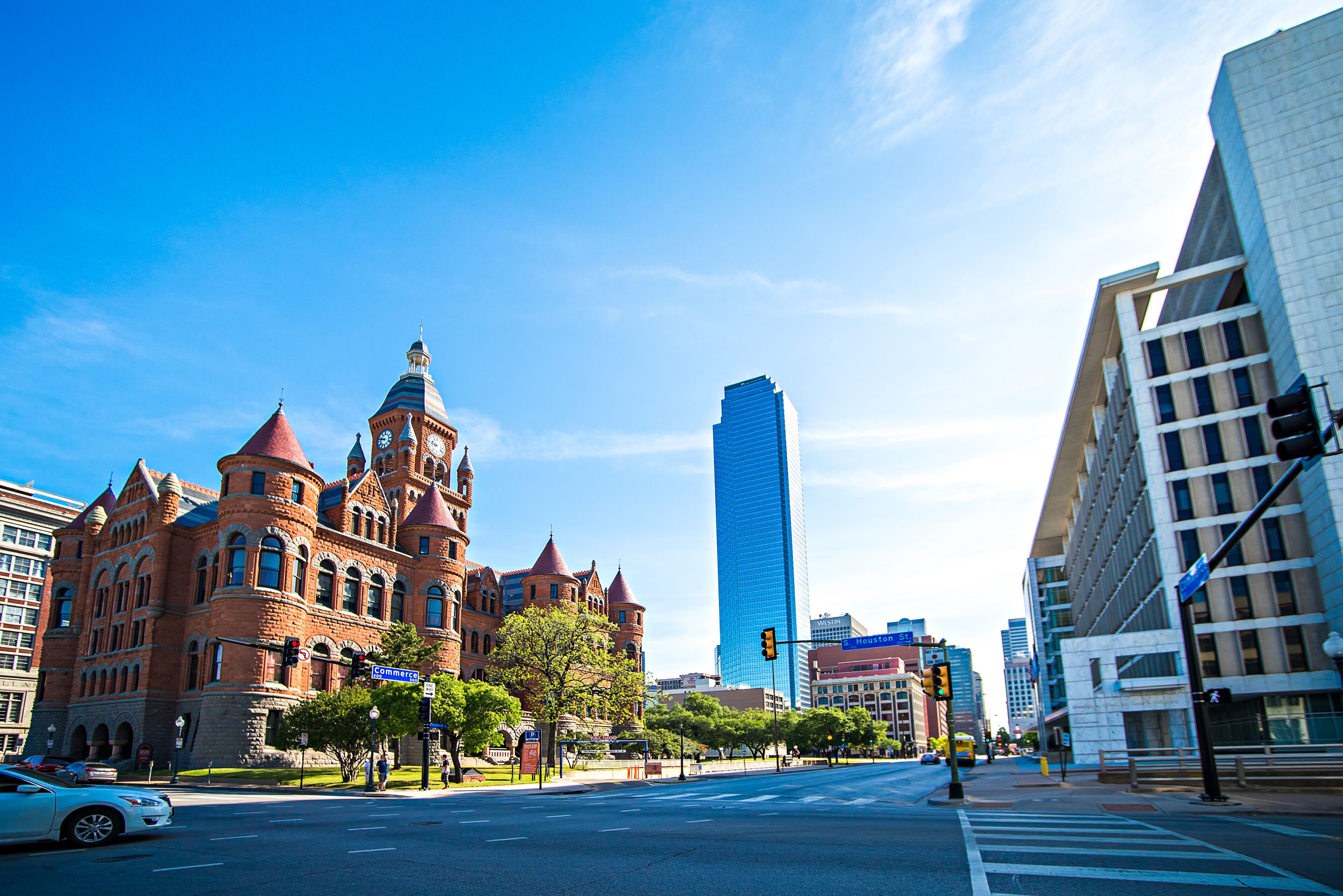Um edifício histórico com tijolos vermelhos e torres, um arranha-céu moderno e uma rua com semáforos.