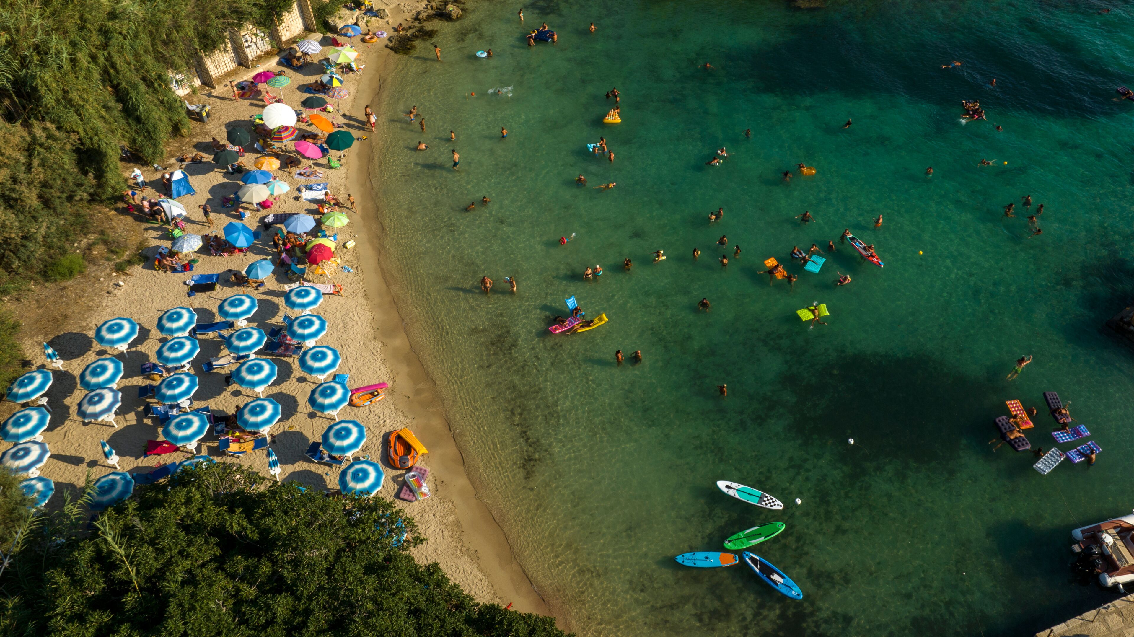 Vista aérea de uma praia com águas turquesa cristalinas, vários guarda-sóis e pessoas nadando e tomando sol.