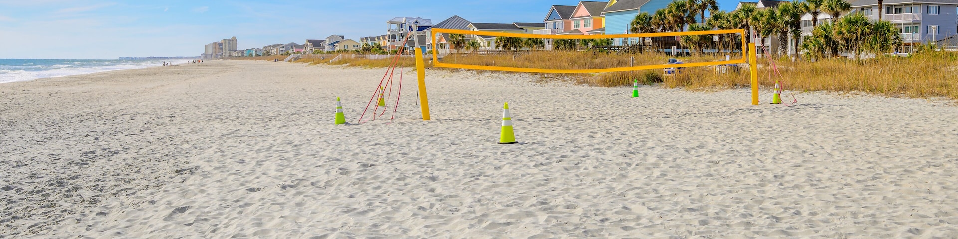 Uma praia arenosa com uma rede de voleibol, cabanas e casas ao fundo.