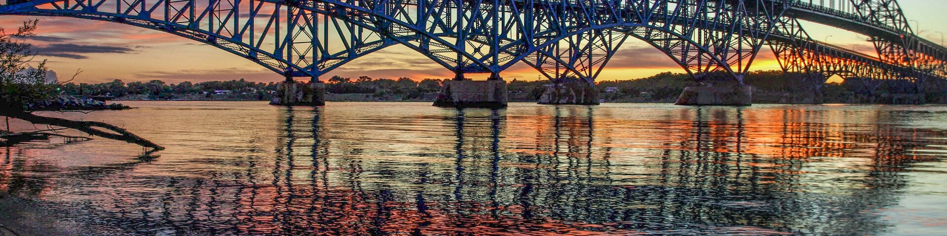 Uma ponte de aço atravessando um rio, com reflexo do pôr do sol.
