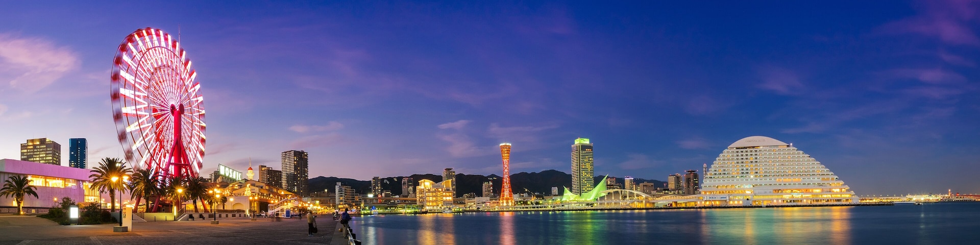 Paisagem urbana à beira-mar com uma roda-gigante vermelha imponente, um grande navio de cruzeiro e edifícios iluminados sob um céu crepuscular.
