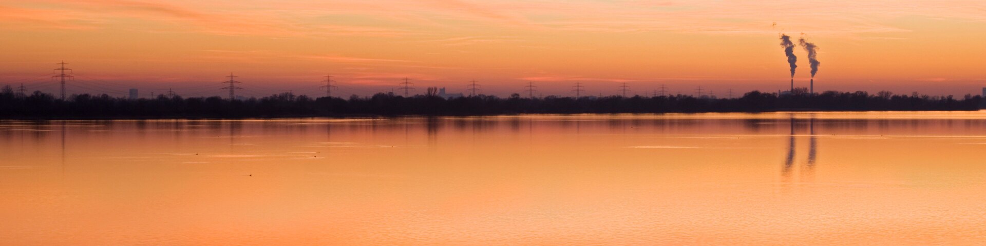 Um lago tranquilo ao pôr do sol, com estruturas industriais ao longe.
