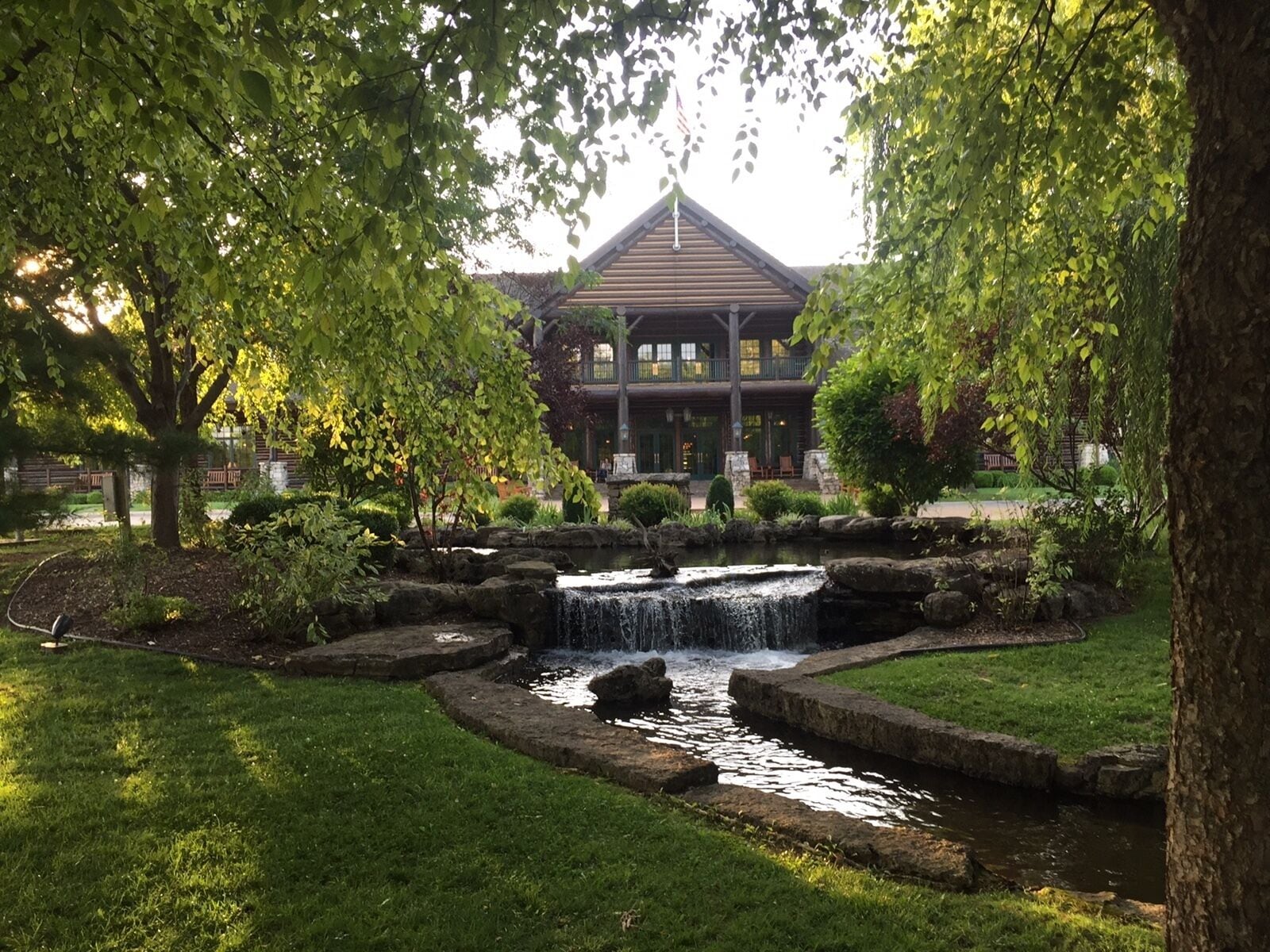 Um jardim tranquilo com uma cascata, vegetação exuberante e um edifício de madeira ao fundo.