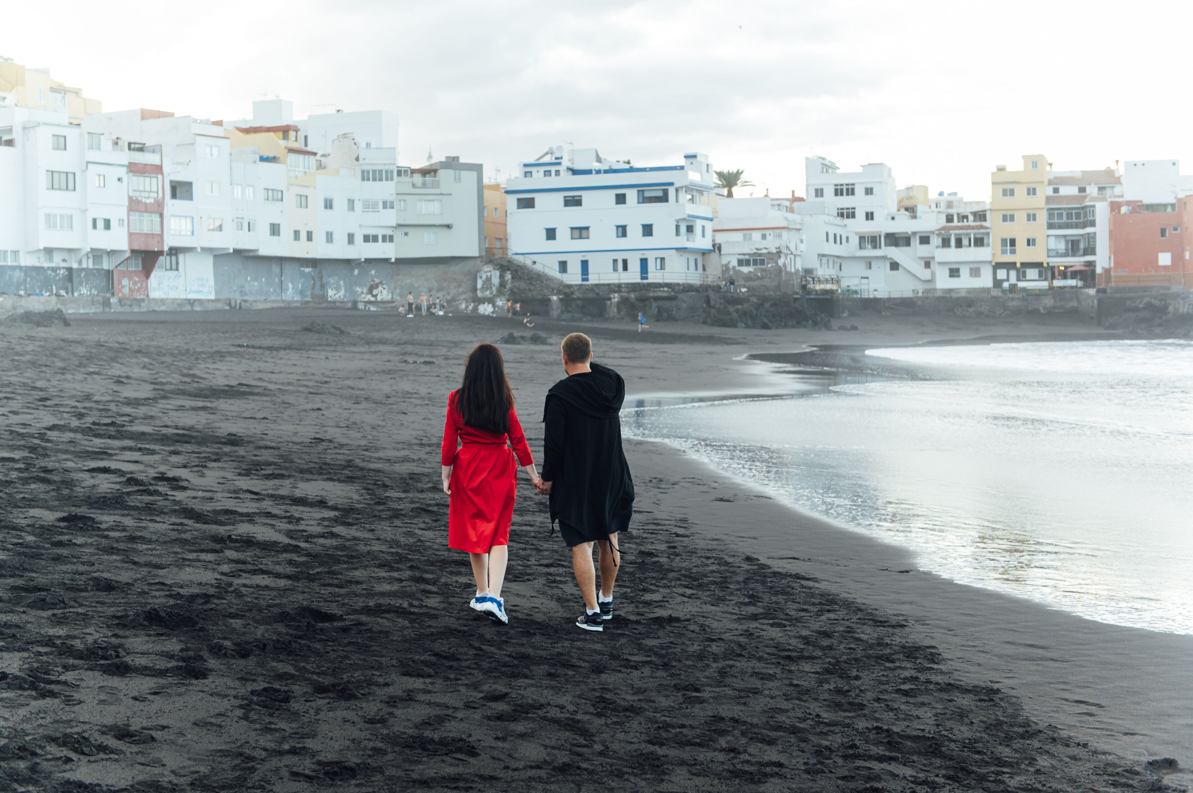 Um casal caminhando numa praia de areia, com edifícios ao fundo.
