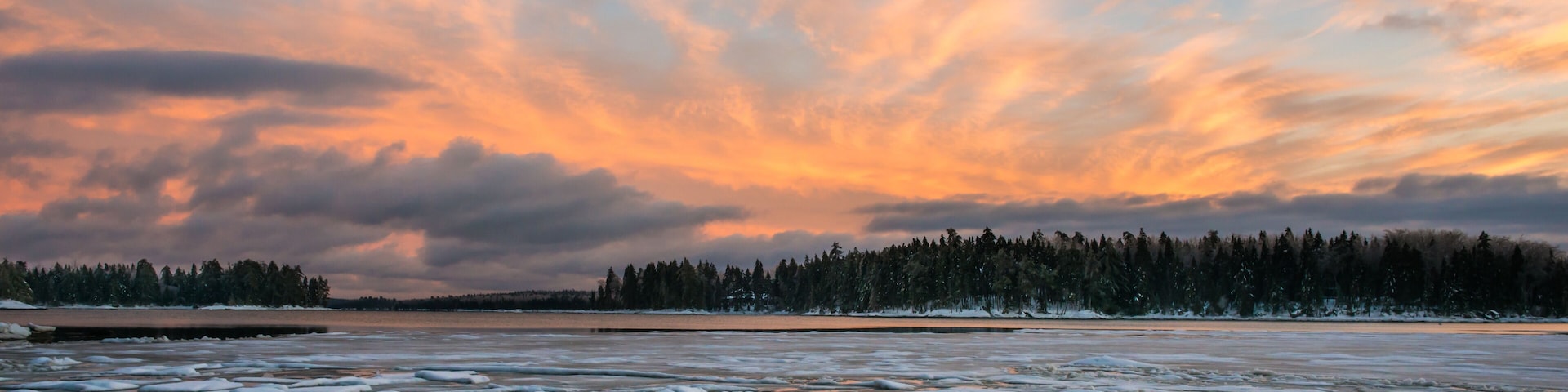 Um lago congelado com icebergs, uma margem arborizada e um céu ao pôr do sol.
