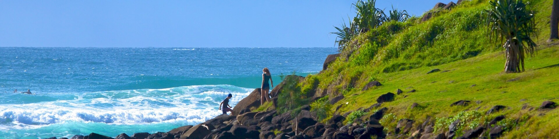 Uma costa rochosa com ondas quebrando nas rochas, uma colina gramada com pessoas em seu topo, e um céu azul límpido.