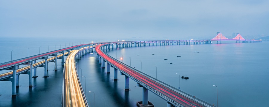 Uma ponte extensa com rastros de luz, mar calmo e estruturas vermelhas ao longe.