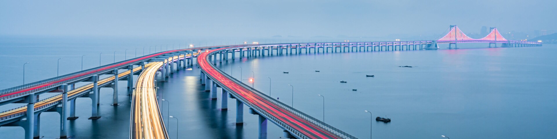 Uma ponte extensa com rastros de luz, mar calmo e estruturas vermelhas ao longe.
