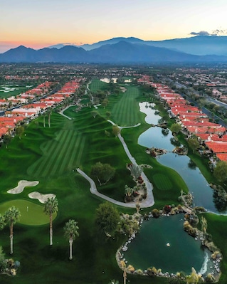 Um campo de golfe com obstáculos de água, armadilhas de areia e montanhas ao fundo.