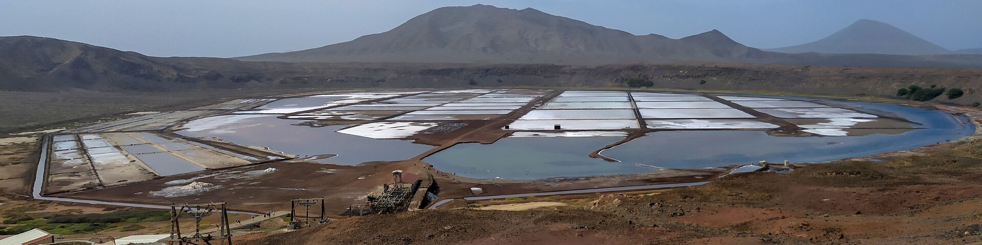 Uma planície salina com piscinas retangulares de água, rodeada por terreno árido e montanhas ao longe.