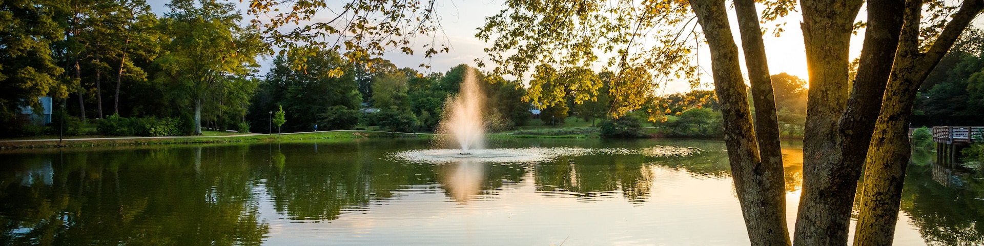 Um lago calmo com uma fonte, rodeado por árvores e áreas ajardinadas.