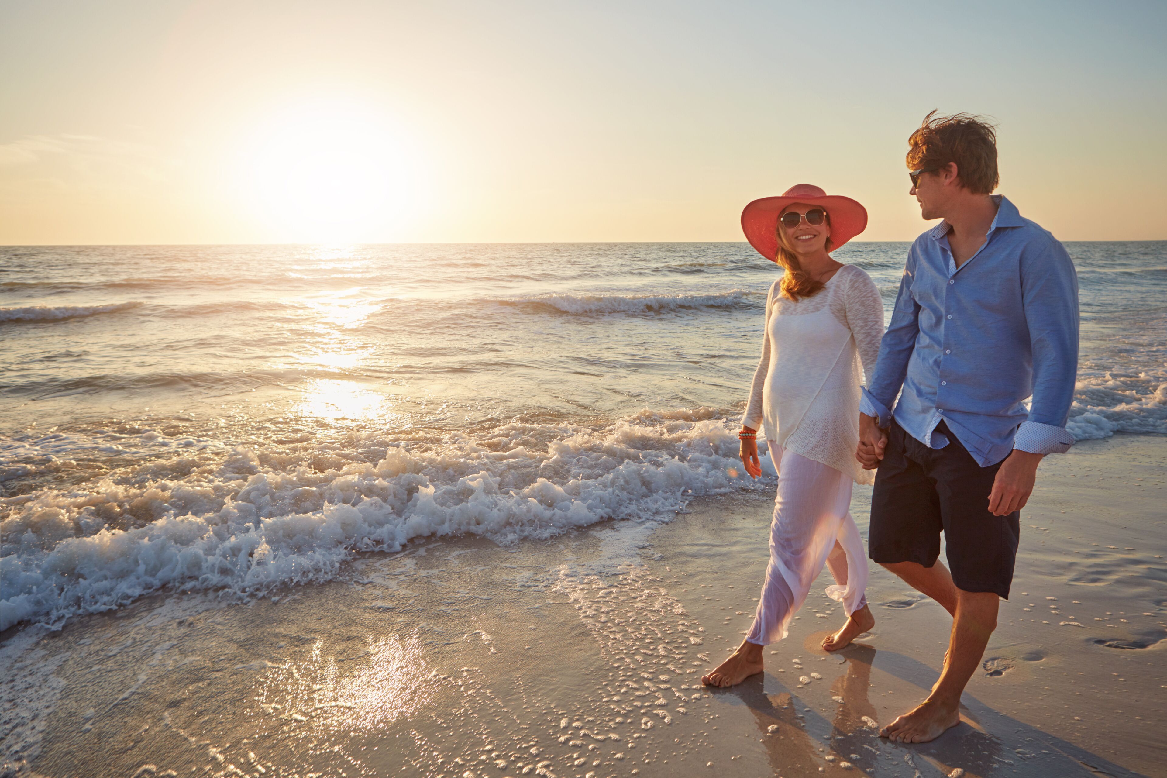 Um casal caminhando na praia com o oceano e um pôr do sol ao fundo.