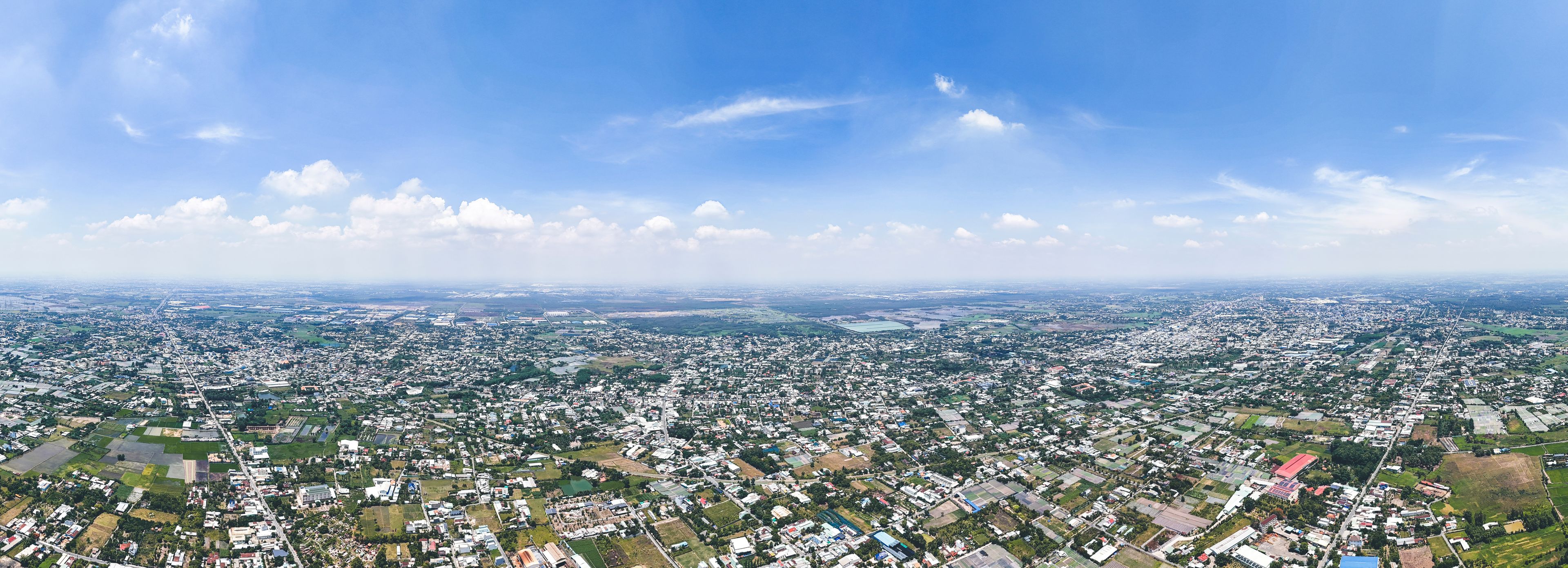 Vista panorâmica de uma cidade densamente povoada, com mistura de edifícios residenciais e comerciais, áreas verdes e céu azul claro com algumas nuvens.