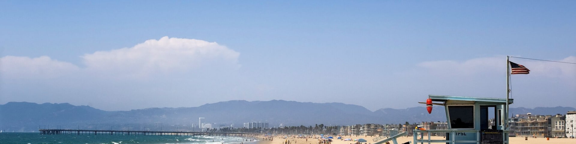 Uma praia com uma torre salva-vidas, ondas do oceano e um cais ao fundo.