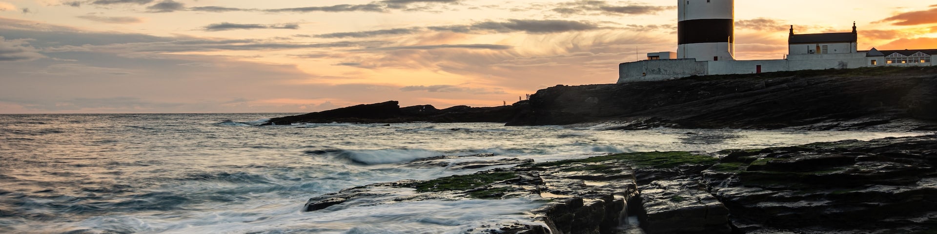 Um farol em um litoral rochoso, com ondas quebrando nas rochas.