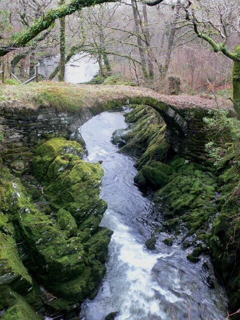 Uma ponte de pedra sobre um rio corrente, com pedras cobertas de musgo e árvores ao fundo.
