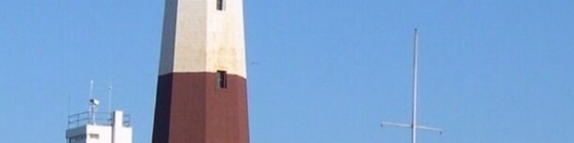 Um farol com torre vermelha e branca, um edifício branco e um mastro de bandeira.