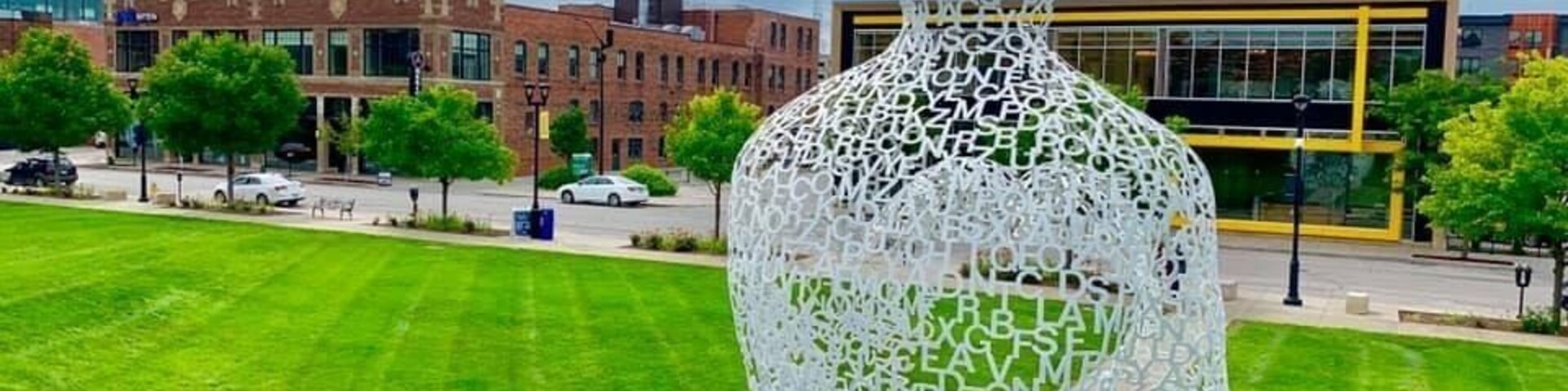 Uma grande escultura branca de uma figura humana em um parque, com edifícios ao fundo.