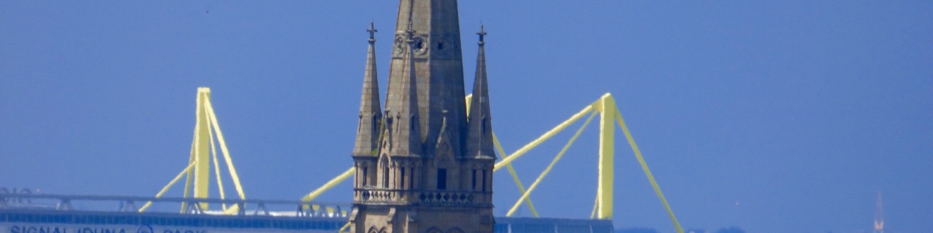 Uma igreja com uma torre alta, uma ponte com cabos amarelos e vegetação em primeiro plano.